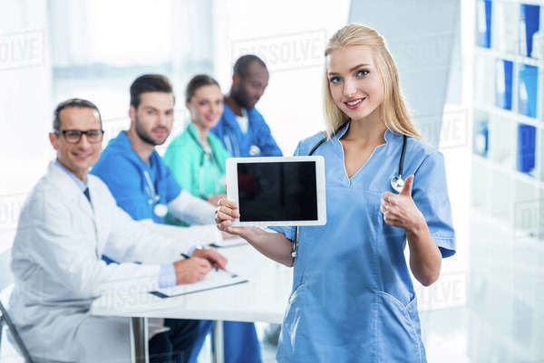 Young nurse holding tablet and showing thumb up with colleagues sitting ...