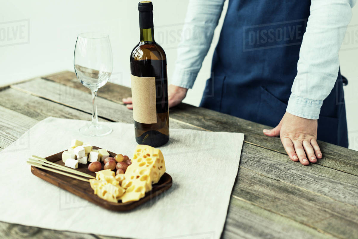 Female sommelier in apron standing at table with wine bottle and food - Stock Photo - Dissolve