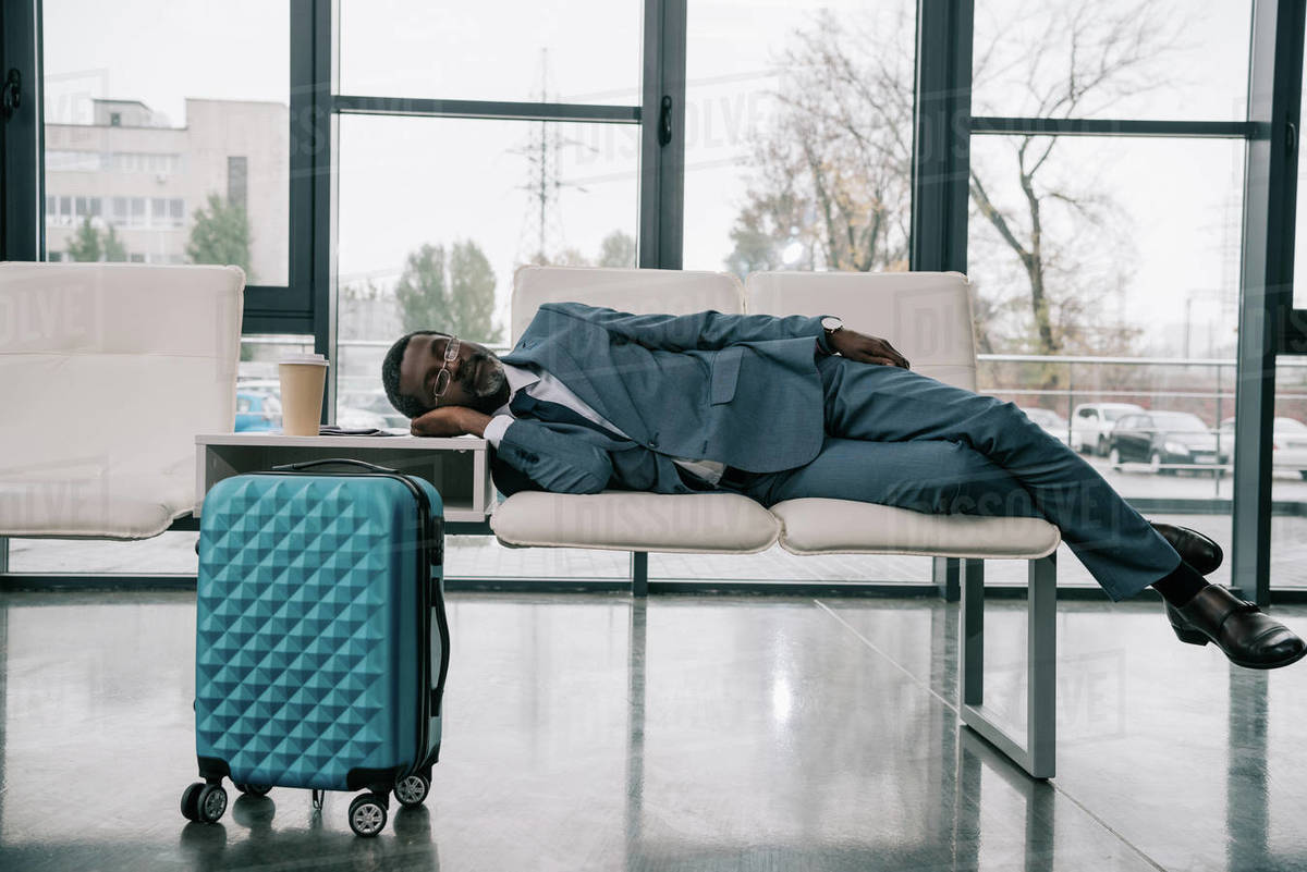 Middle aged African American businessman napping on chairs at airport ...