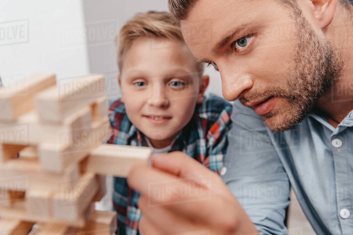 Father pulling a piece out of blocks wood tower while son is watching ...