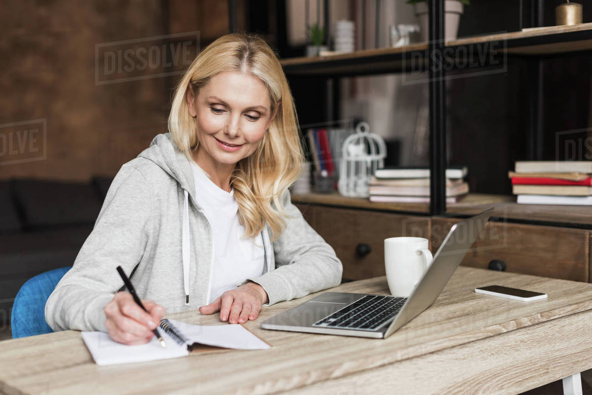 smiling mature woman taking notes and using laptop at home - Royalty ...