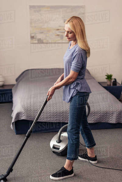 side view of beautiful blonde woman using vacuum cleaner at home ...