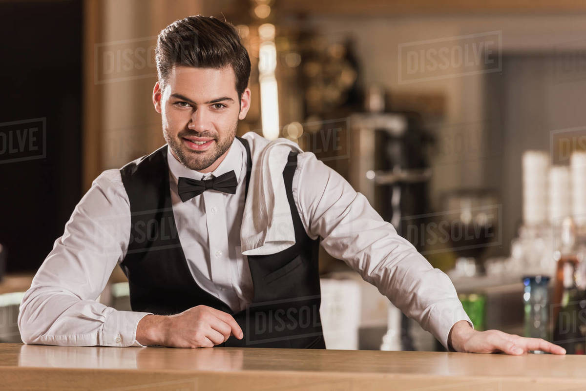 Handsome barman leaning on bar counter and looking at the camera ...