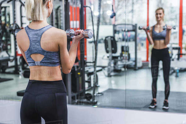 Athletic young woman working out with dumbbells in front of mirror at ...