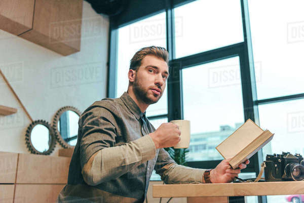 Side view of man with cup of coffee and book in hands sitting at table ...