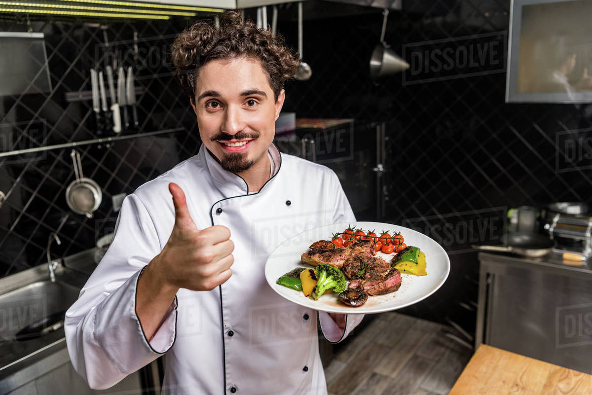 Happy chef showing thumb up and holding cooked vegetables with meat ...