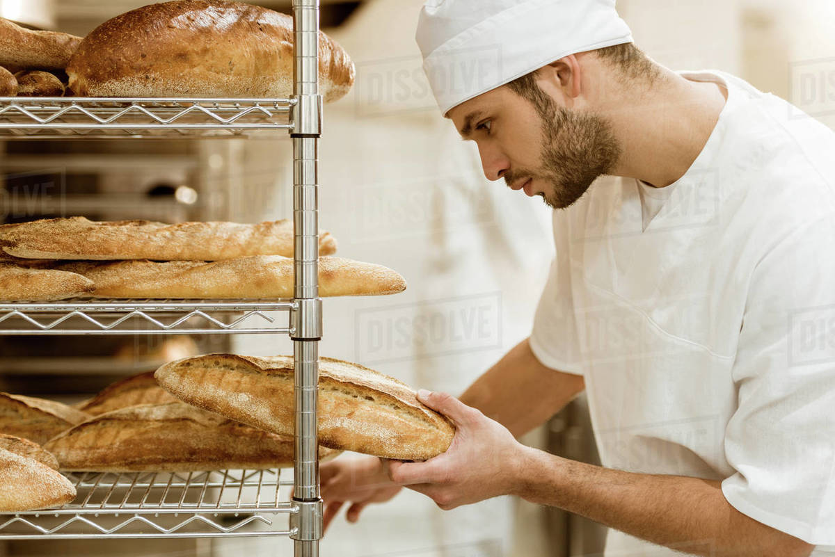 Side view of handsome baker putting fresh bread on shelf at baking ...