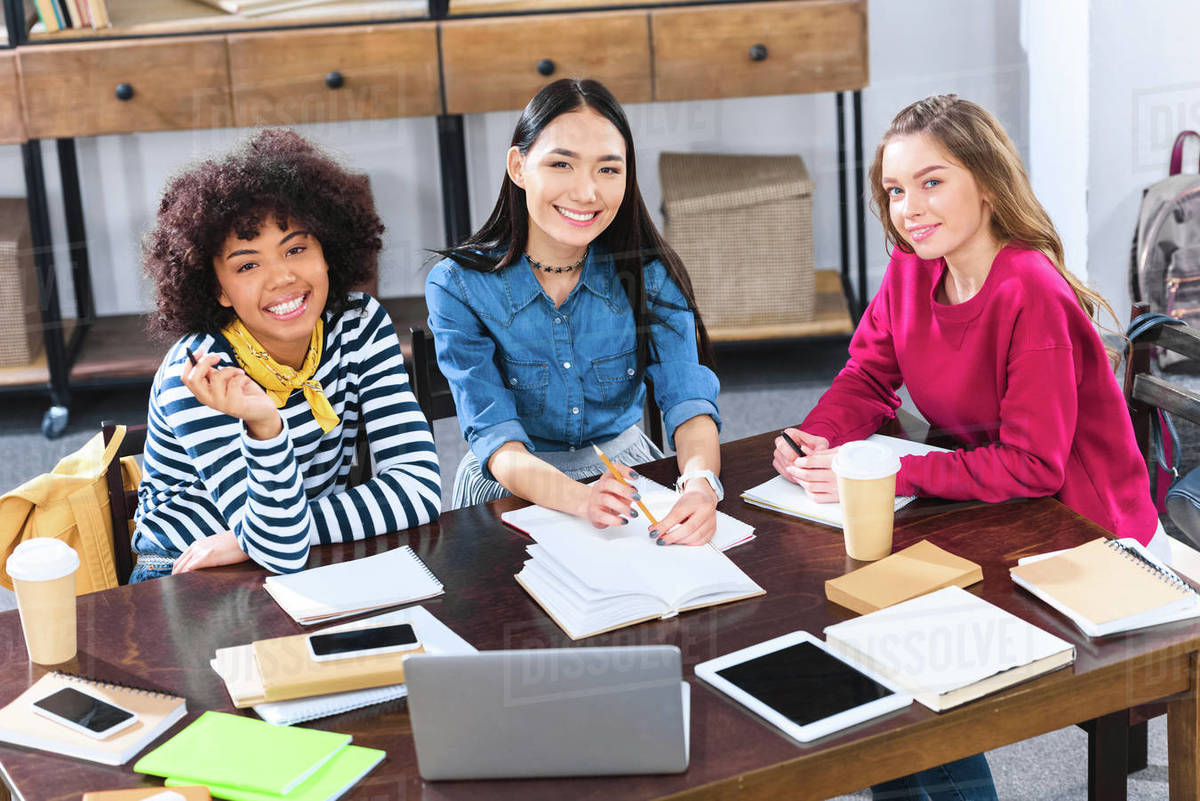 Smiling multiracial students looking at camera while doing homework ...