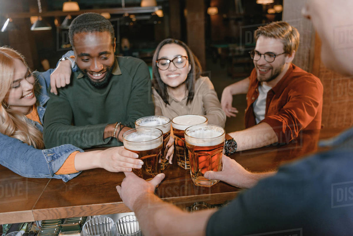 Cropped image of barman giving beer to multicultural company of friends ...