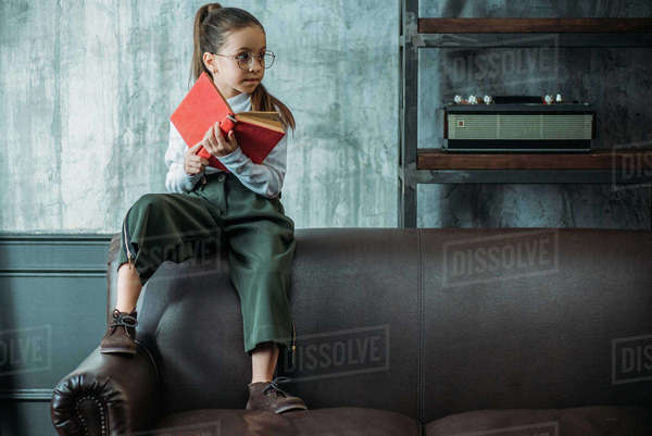 Thoughtful little child with book sitting on couch in loft apartments ...