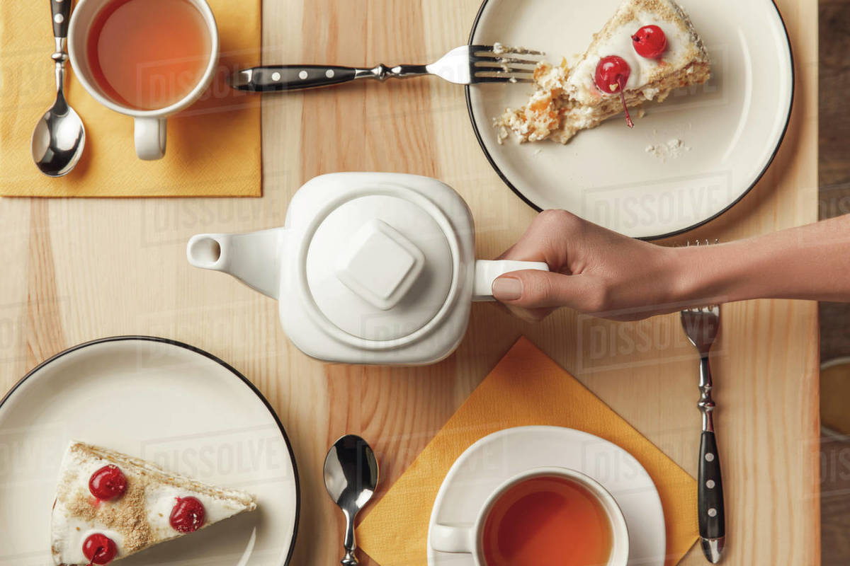 Top view of person holding teapot and tea set with pastries on table ...