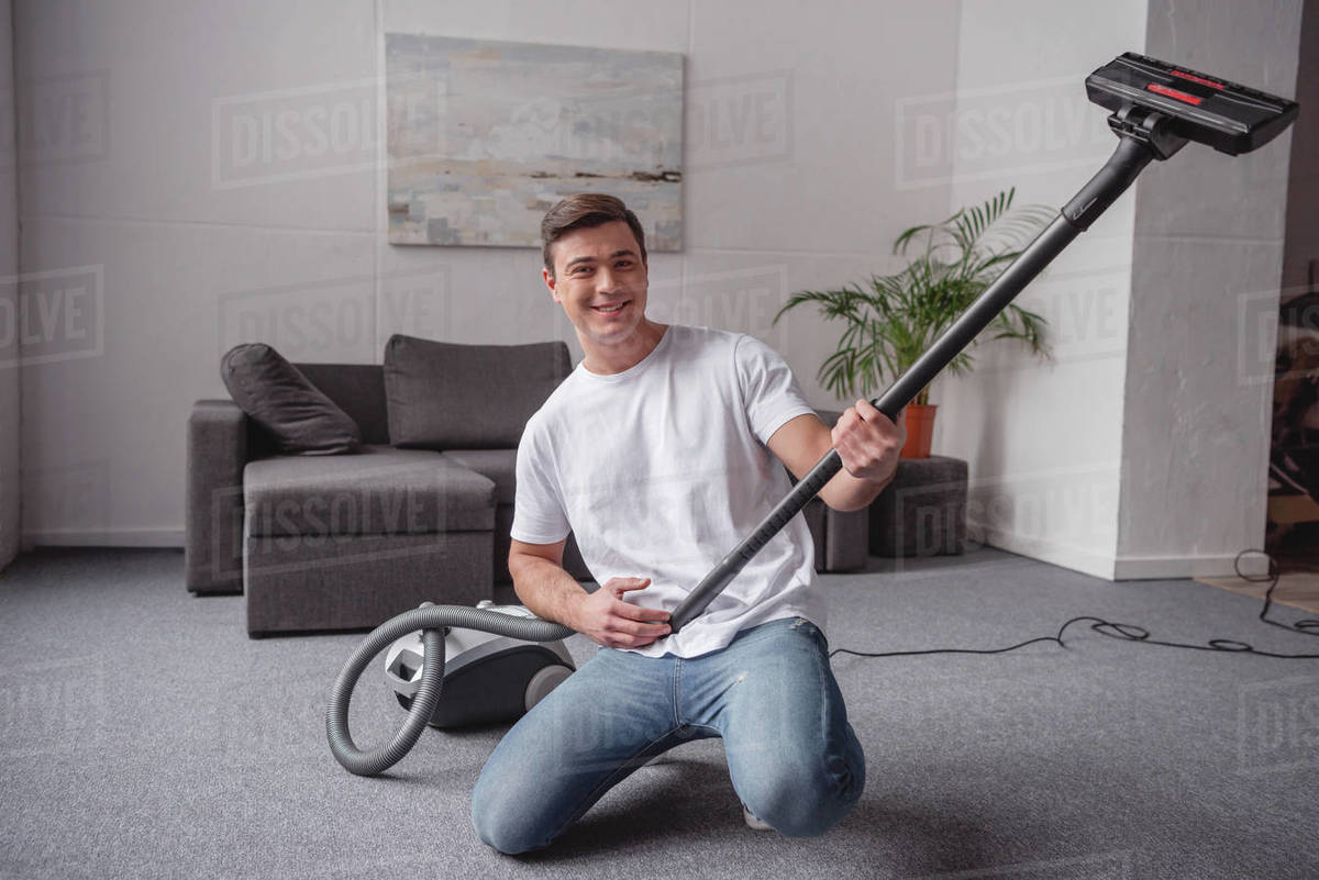 Handsome man having fun with vacuum cleaner in living room and looking ...