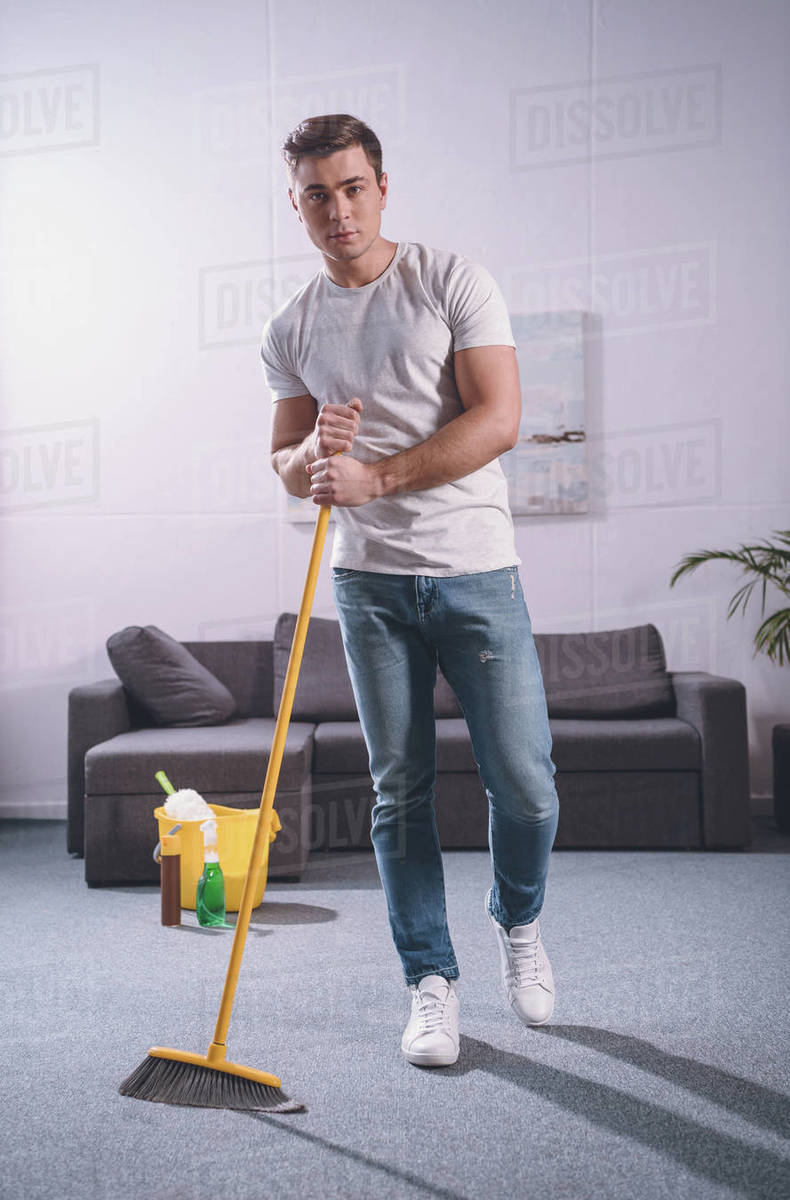Handsome man sweeping living room with broom and looking at camera ...