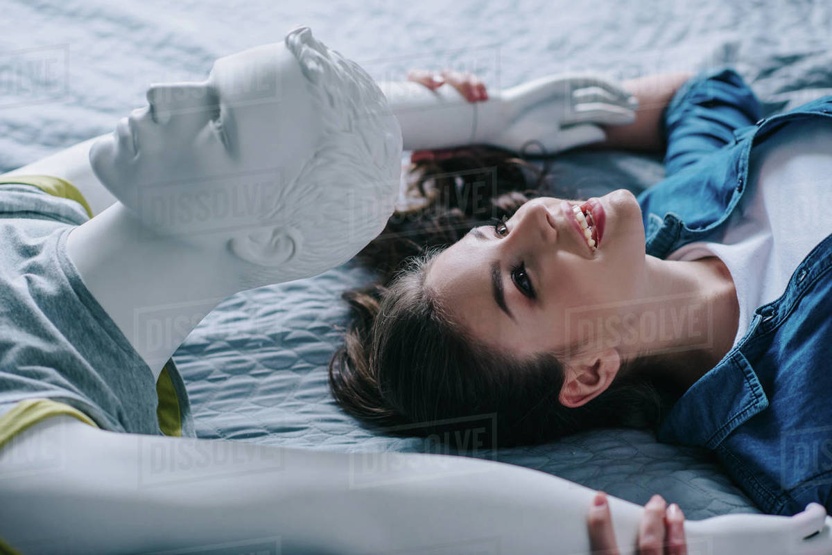 Side view of smiling woman and mannequin lying on bed, loneliness ...