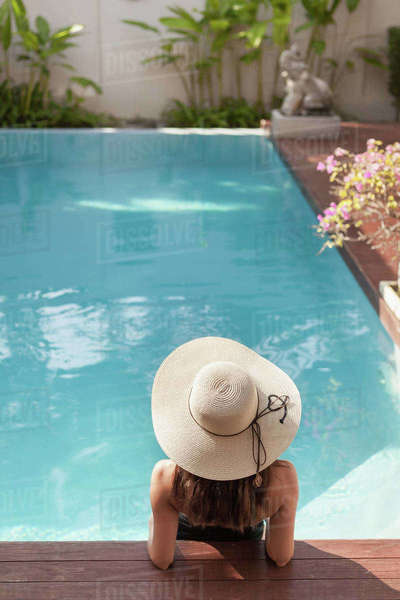Back view of young woman in swimsuit and hat sitting in pool of hotel ...