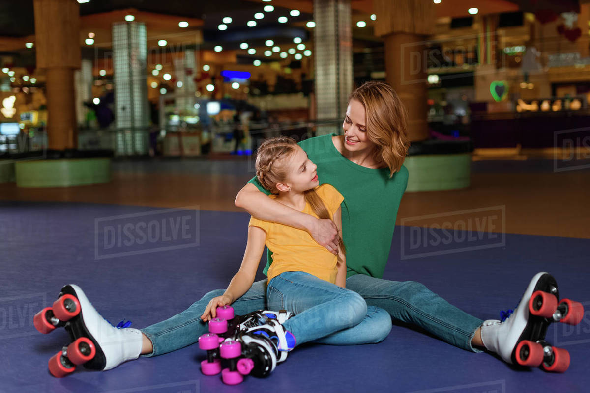 Smiling mother and little daughter sitting on roller rink together ...