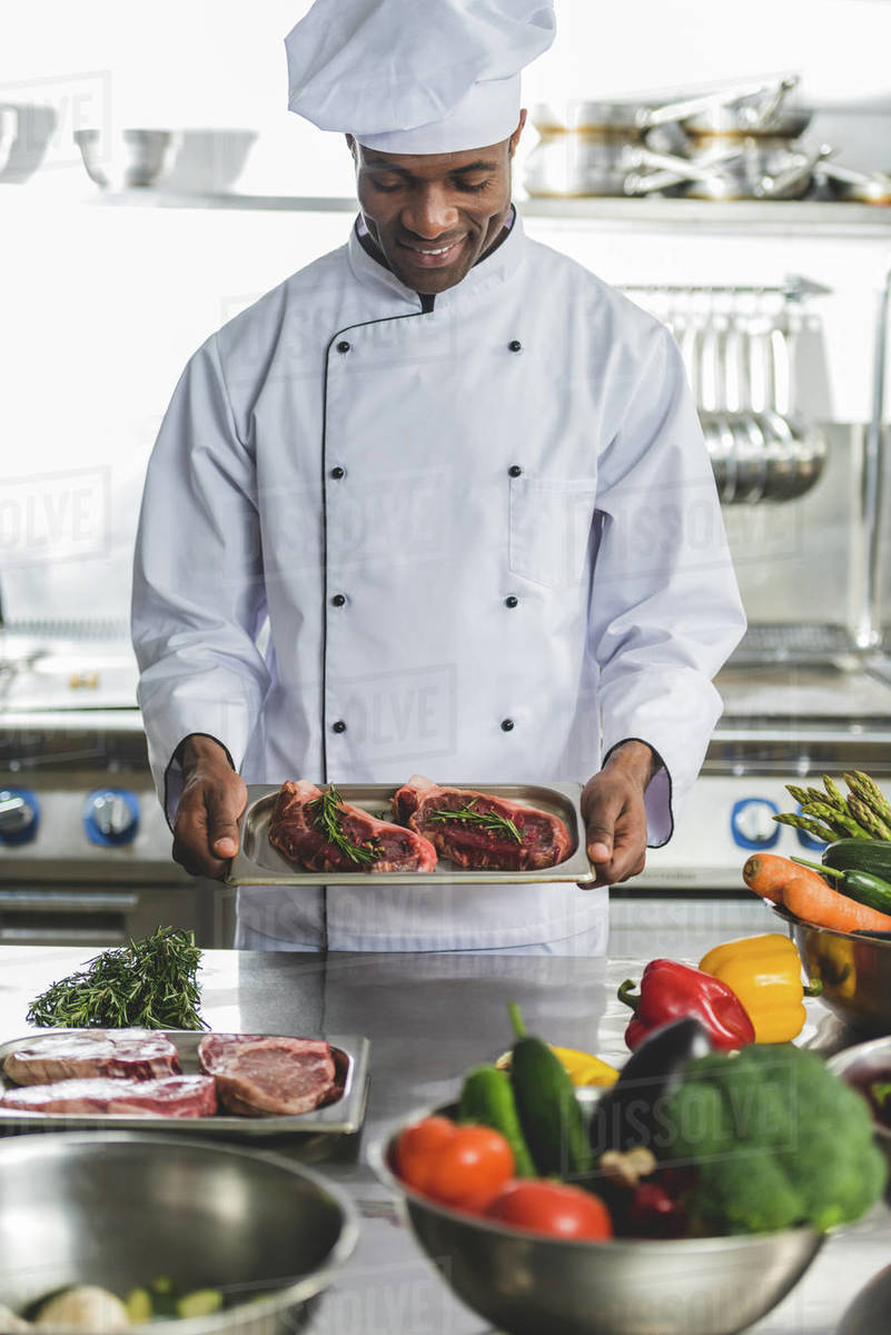 Smiling handsome African American chef holding tray with uncooked meat ...