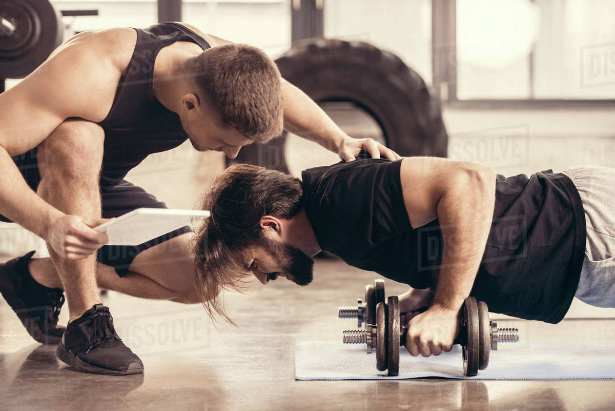 Side view of trainer pushing sportsman doing push ups on dumbbells in ...