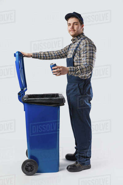 Side view of young cleaner in uniform with can standing at trash bin ...