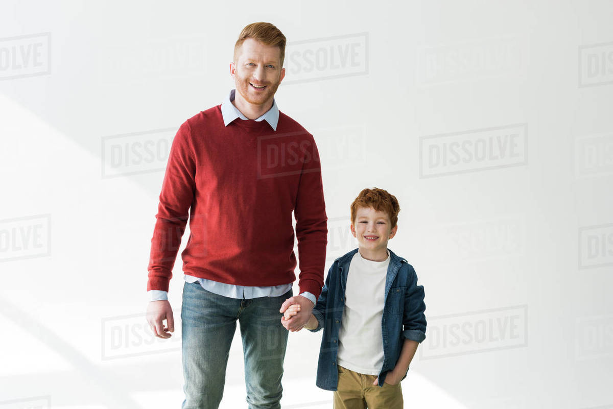 Happy redhead father and son holding hands and smiling at camera on ...