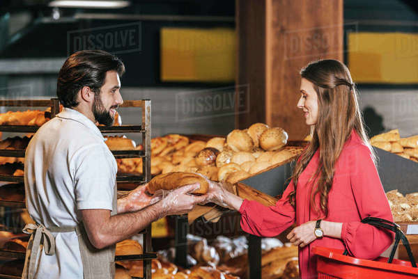 Side view of shop assistant giving loaf of bread to woman with shopping ...