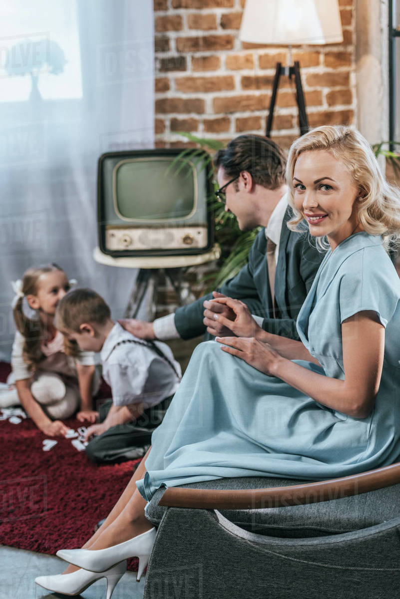 Happy 50s style woman smiling at camera while sitting on sofa with ...