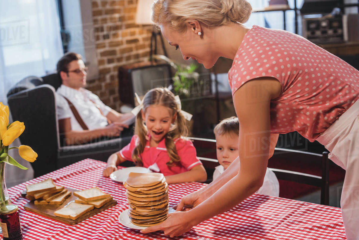 Happy kids looking at smiling mother putting pancakes on table, retro ...