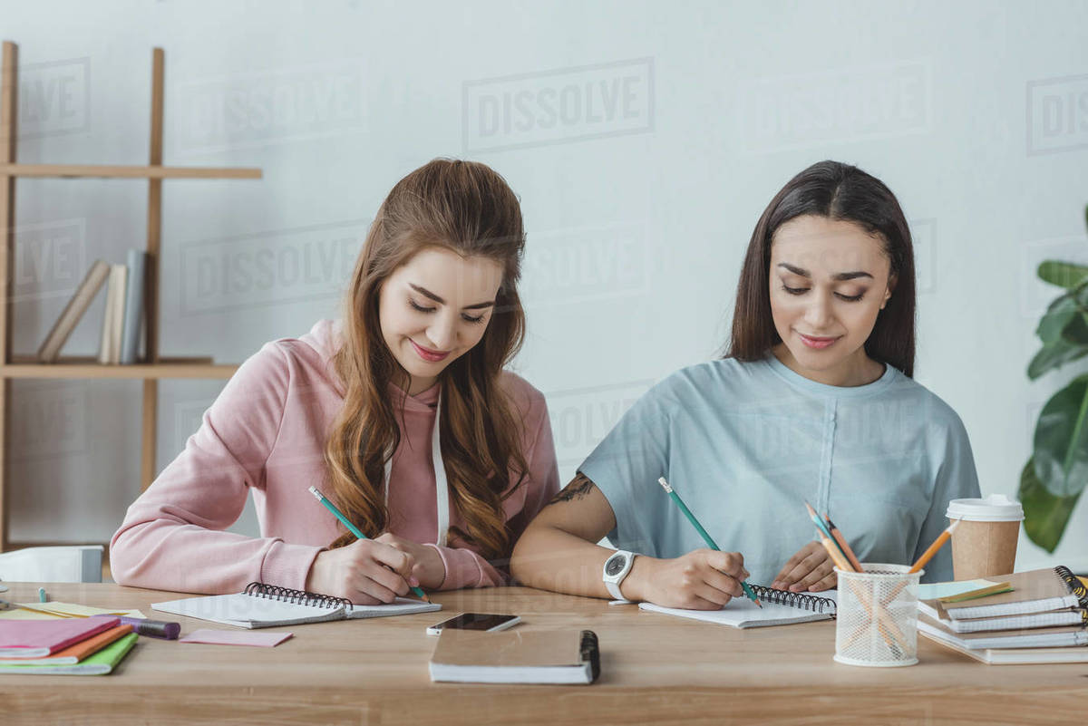 Multiethnic girls studying together and writing in copybooks at table ...