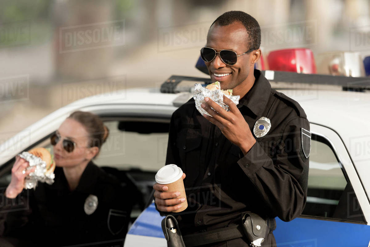African american policeman with paper cup of coffee eating burger while ...
