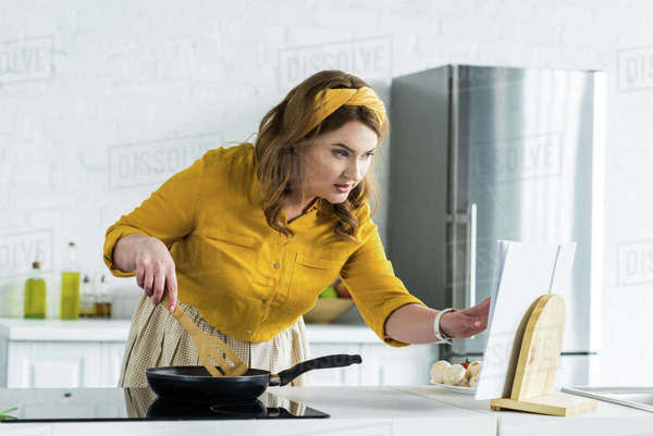 Beautiful woman looking at recipe book and frying vegetables on frying ...