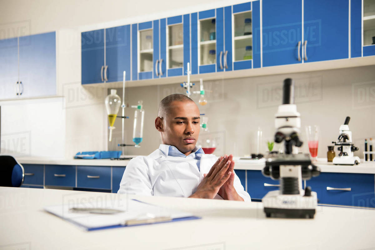 Young thoughtful technician in lab coat sitting in laboratory with ...