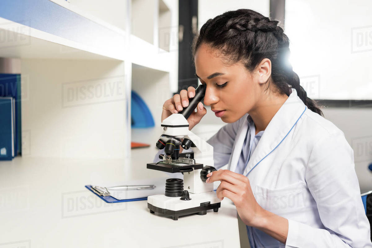 Young lab technician in white coat analyzing samples using microscope ...