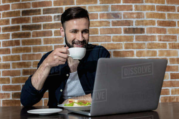 Bearded young man using laptop while eating sandwich and drinking ...