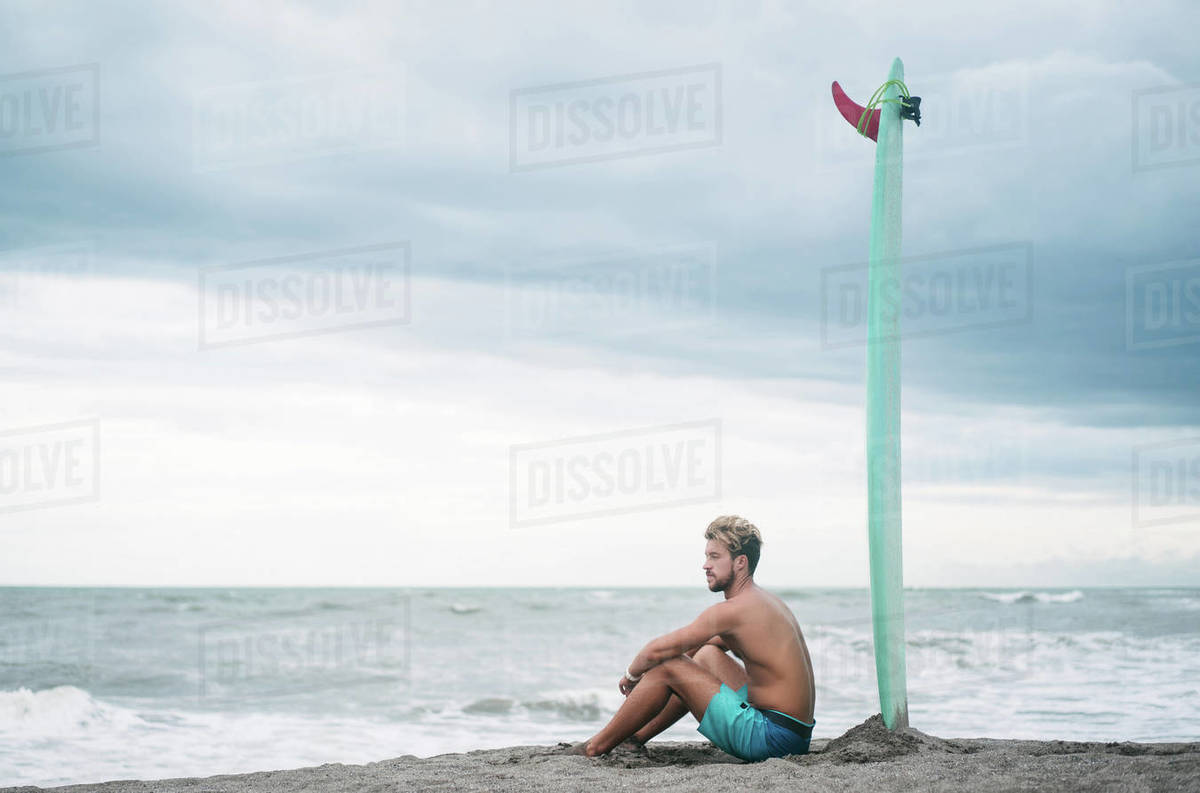 Side view of surfer sitting with surfboard on sandy beach in Bali ...