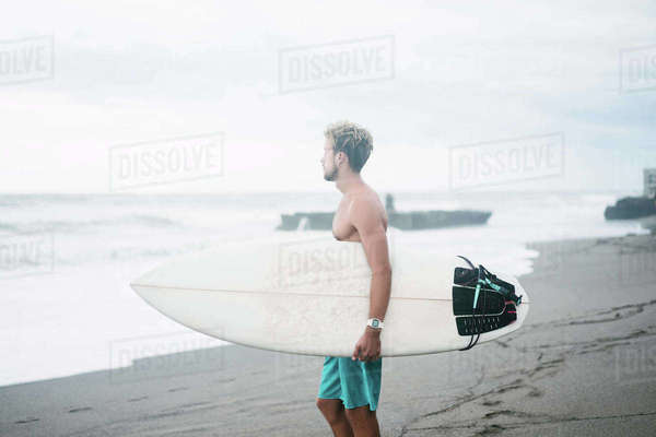 Side view of handsome surfer standing with surfboard in Bali, Indonesia ...