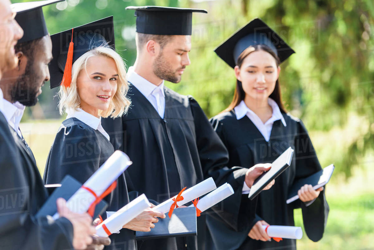 Successful graduated students in capes and hats holding rolled diplomas ...