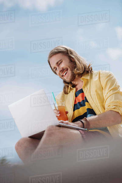 Low angle view of smiling young man holding cocktail and using laptop ...