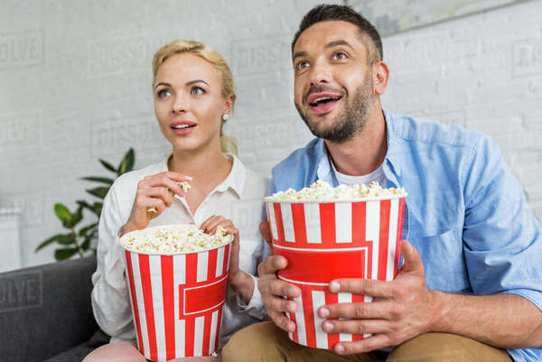 Excited couple eating popcorn and looking away at home - Royalty-free ...