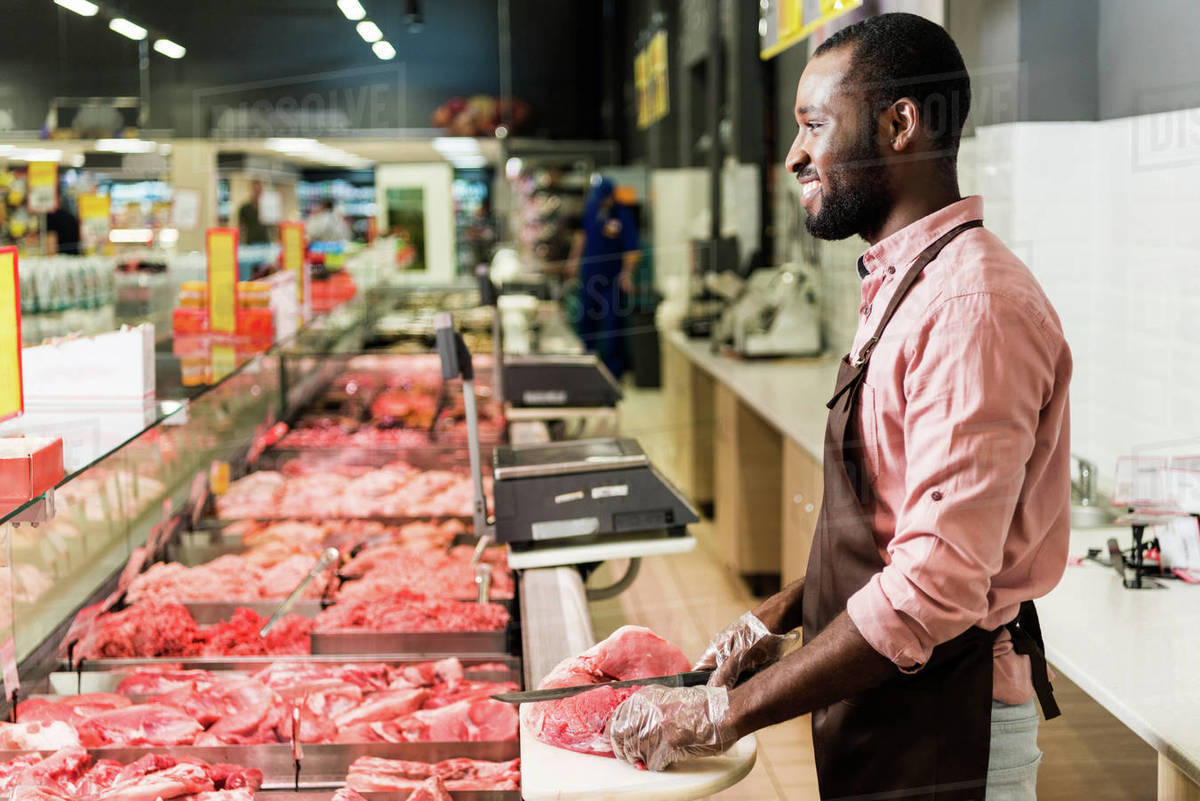 Side view of smiling african american male butcher in apron cutting raw ...