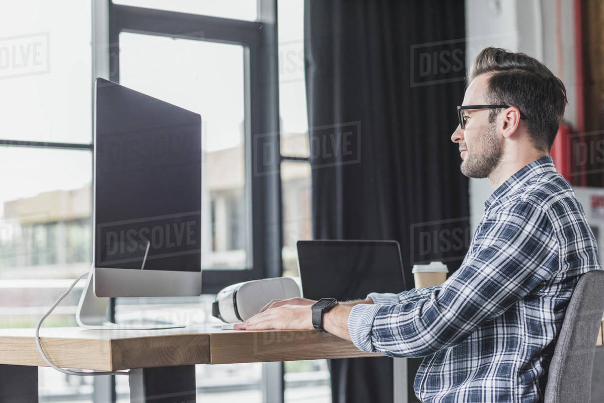 handsome smiling young programmer in eyeglasses working with desktop computer and laptop - Stock ...
