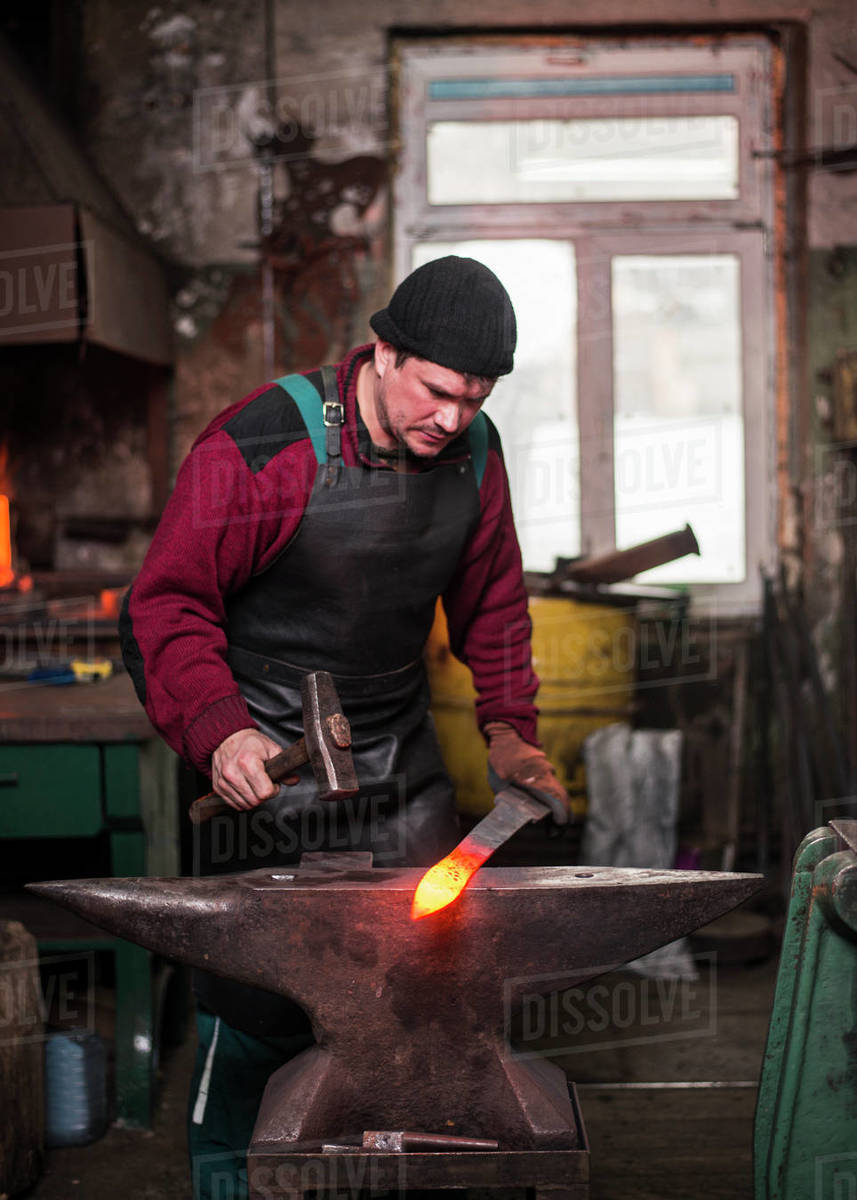 Blacksmith working with a hammer with hot iron piece making a sword ...