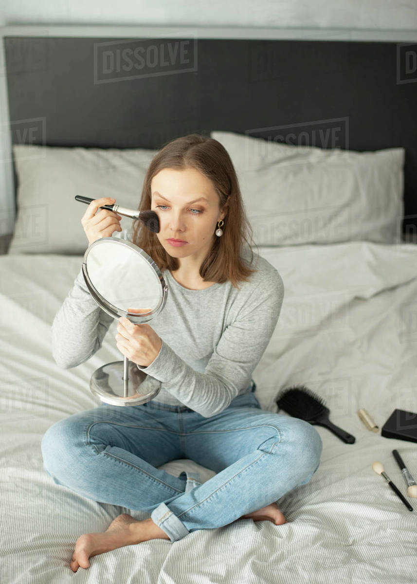 Woman sitting on bed applying makeup on her face - Stock Photo - Dissolve