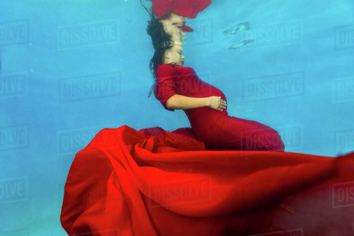 Beautiful pregnant woman in red dress floating underwater with hands on ...