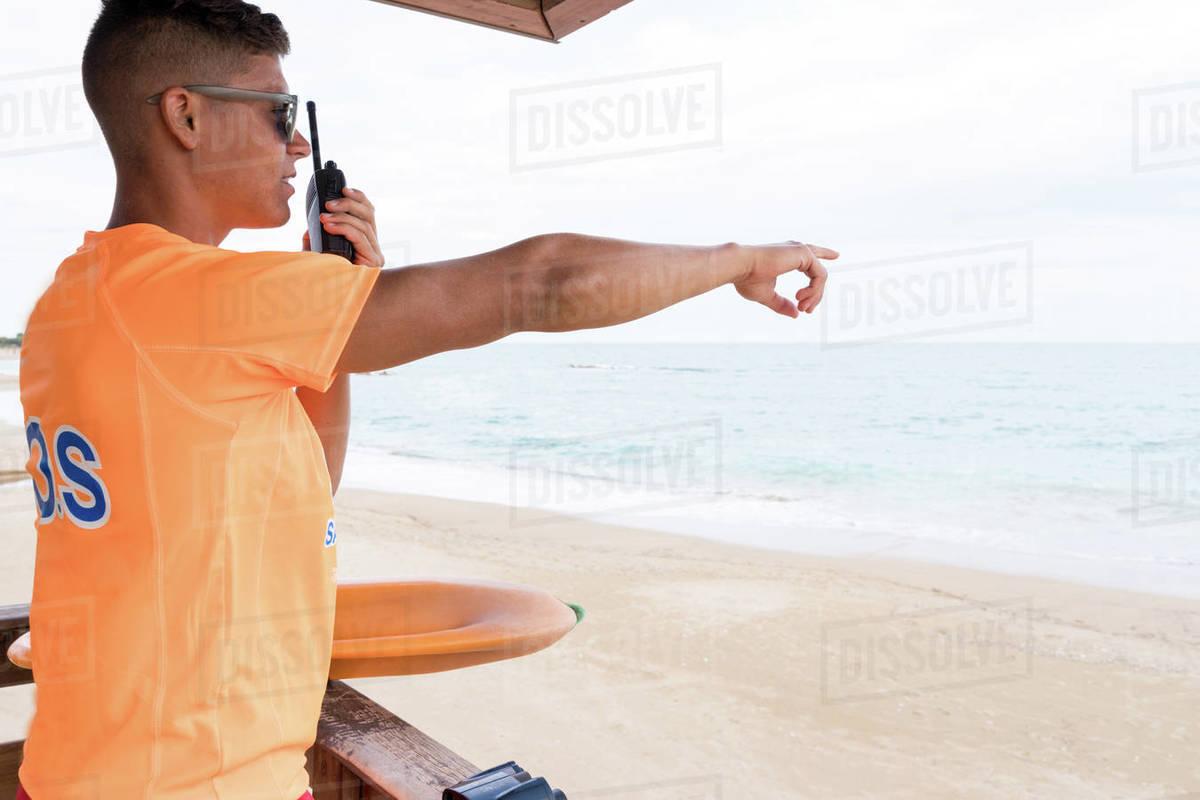 Side view of crop male lifeguard in bright wear pointing with finger at ...