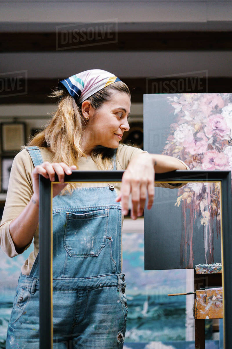 Creative young female artist in denim overall leaning on empty frame in ...