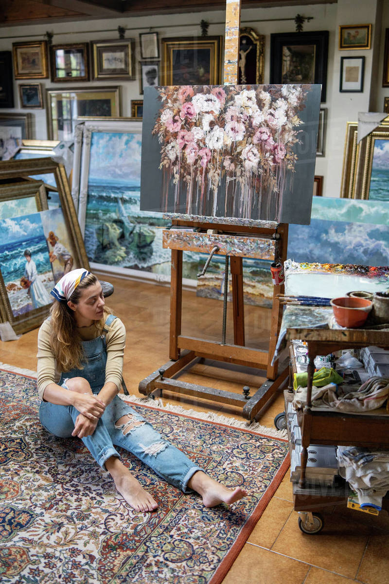 Female artist in denim overall sitting on the floor near easel in ...
