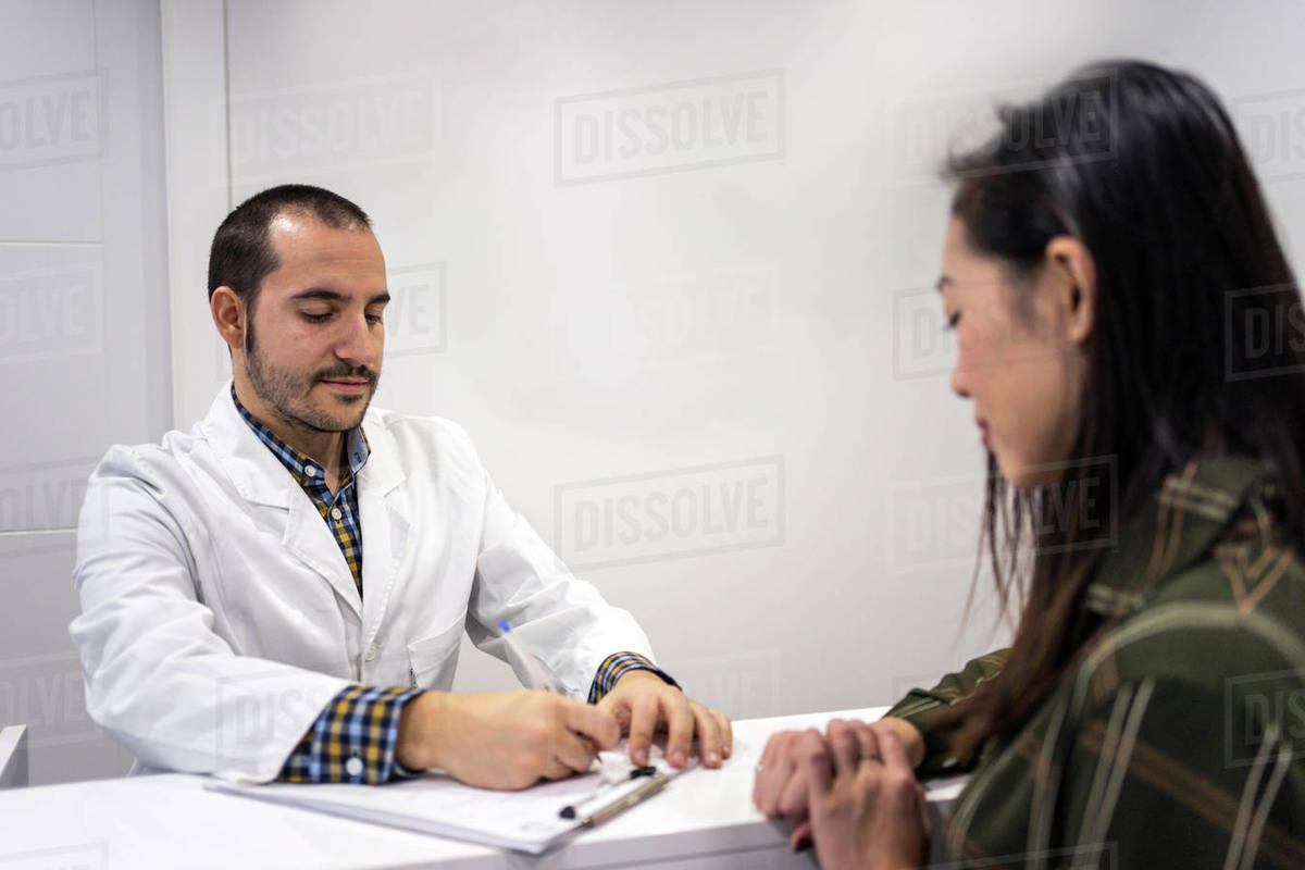 Attentive male doctor filling out paper form while standing with Asian ...