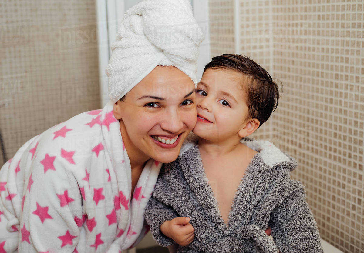 Cheerful woman with towel turban cuddling little child in bathrobe ...