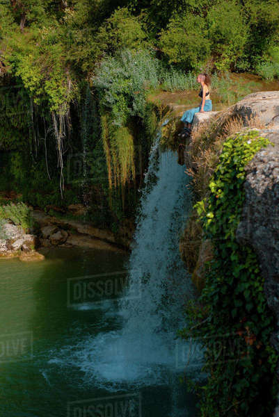 Side view of young female sitting on cliff near waterfall with fresh ...