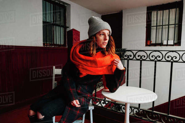 Thoughtful young lady in winter wear looking away and sitting on chair ...